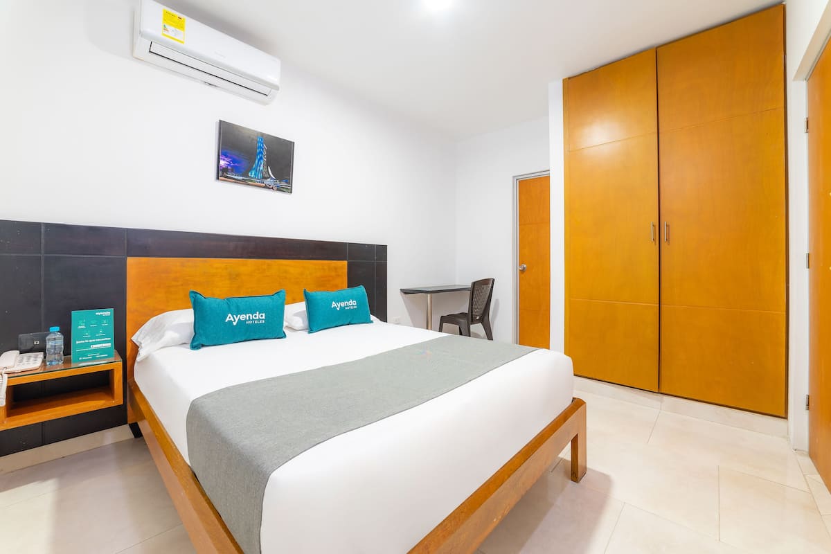 Quarto de hotel com cama, escrivaninha, ar condicionado, um quadro na parede e um guarda-roupa.