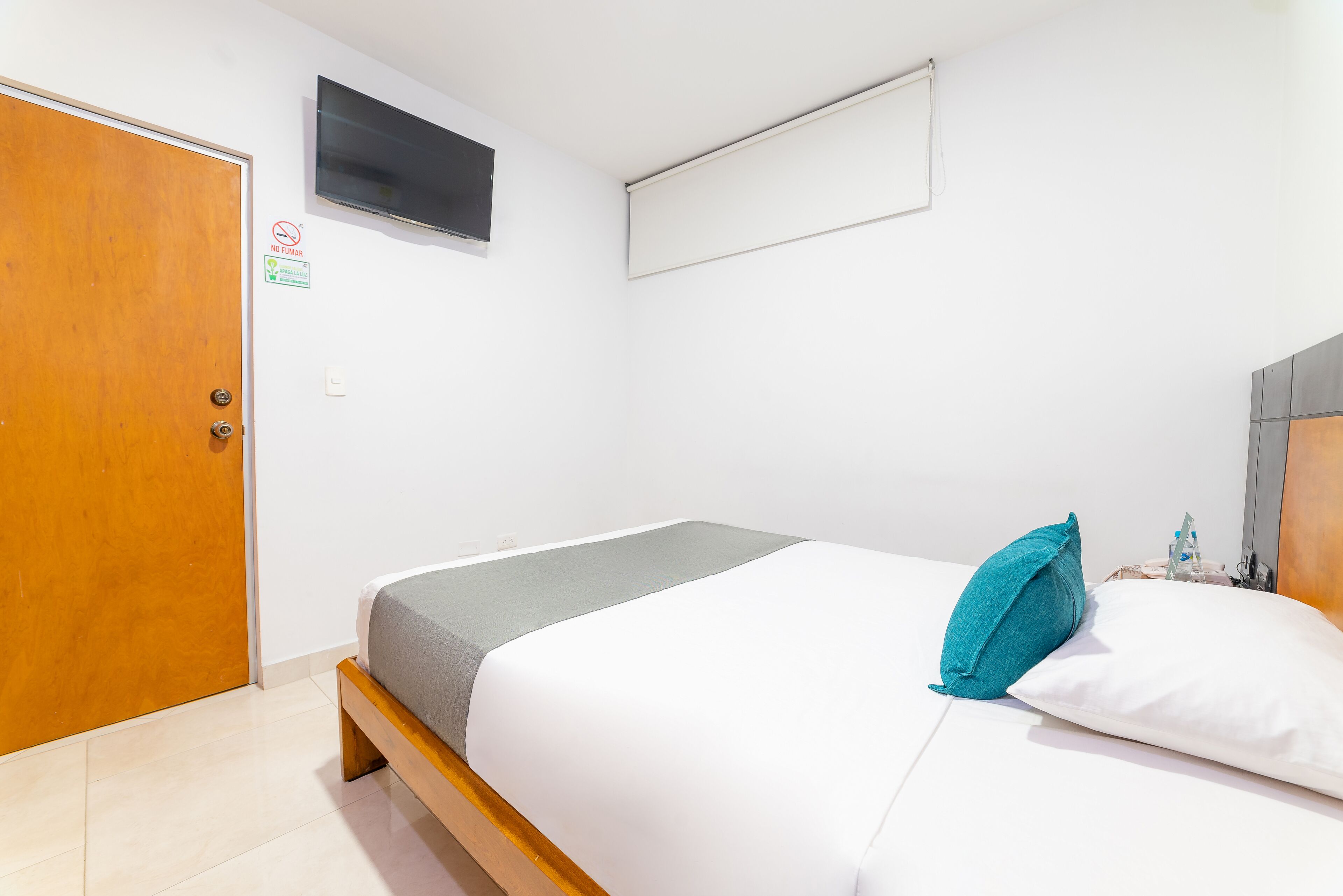 Quarto com cama, porta de madeira, televisão fixada na parede e placa indicando proibido fumar.