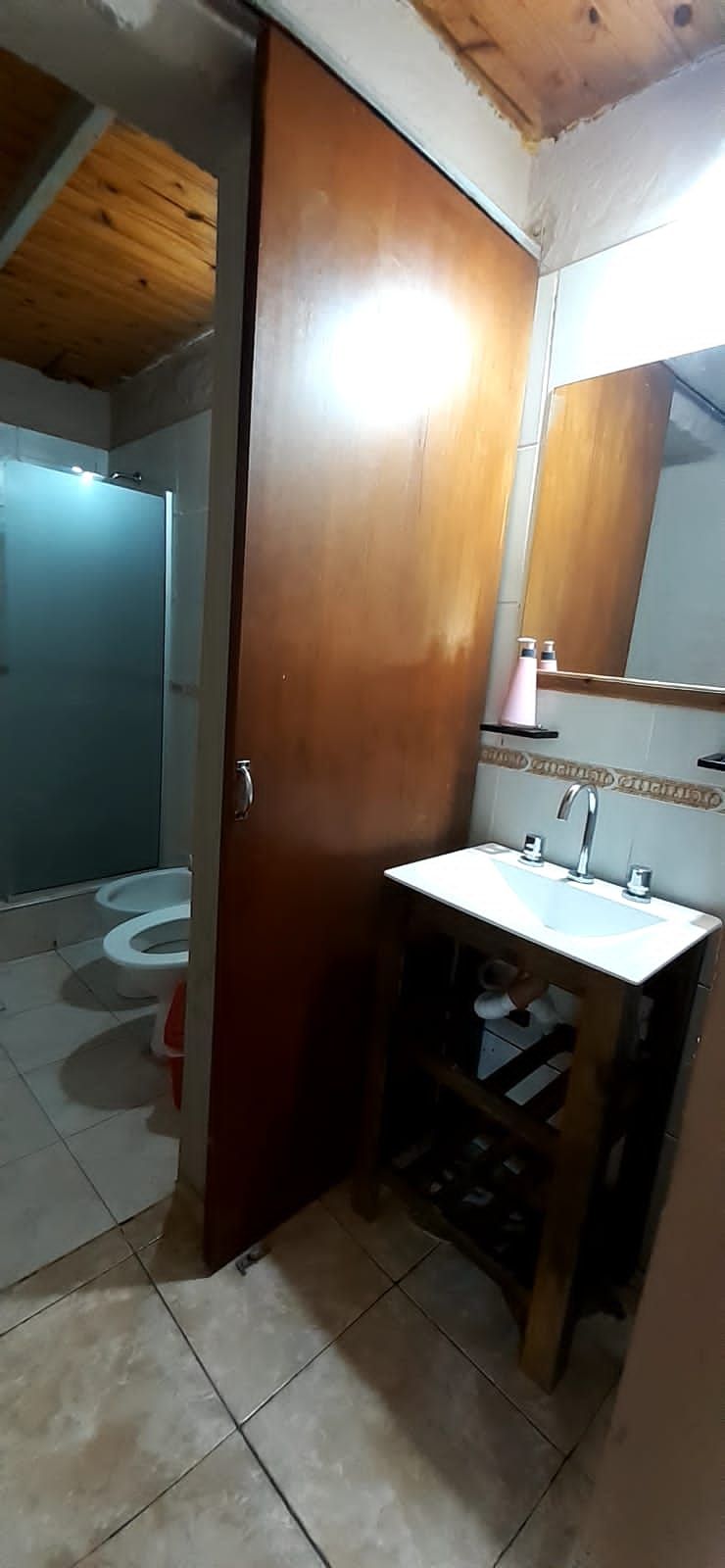 Casa de banho com porta de madeira, pia branca, espelho e zona de duche.