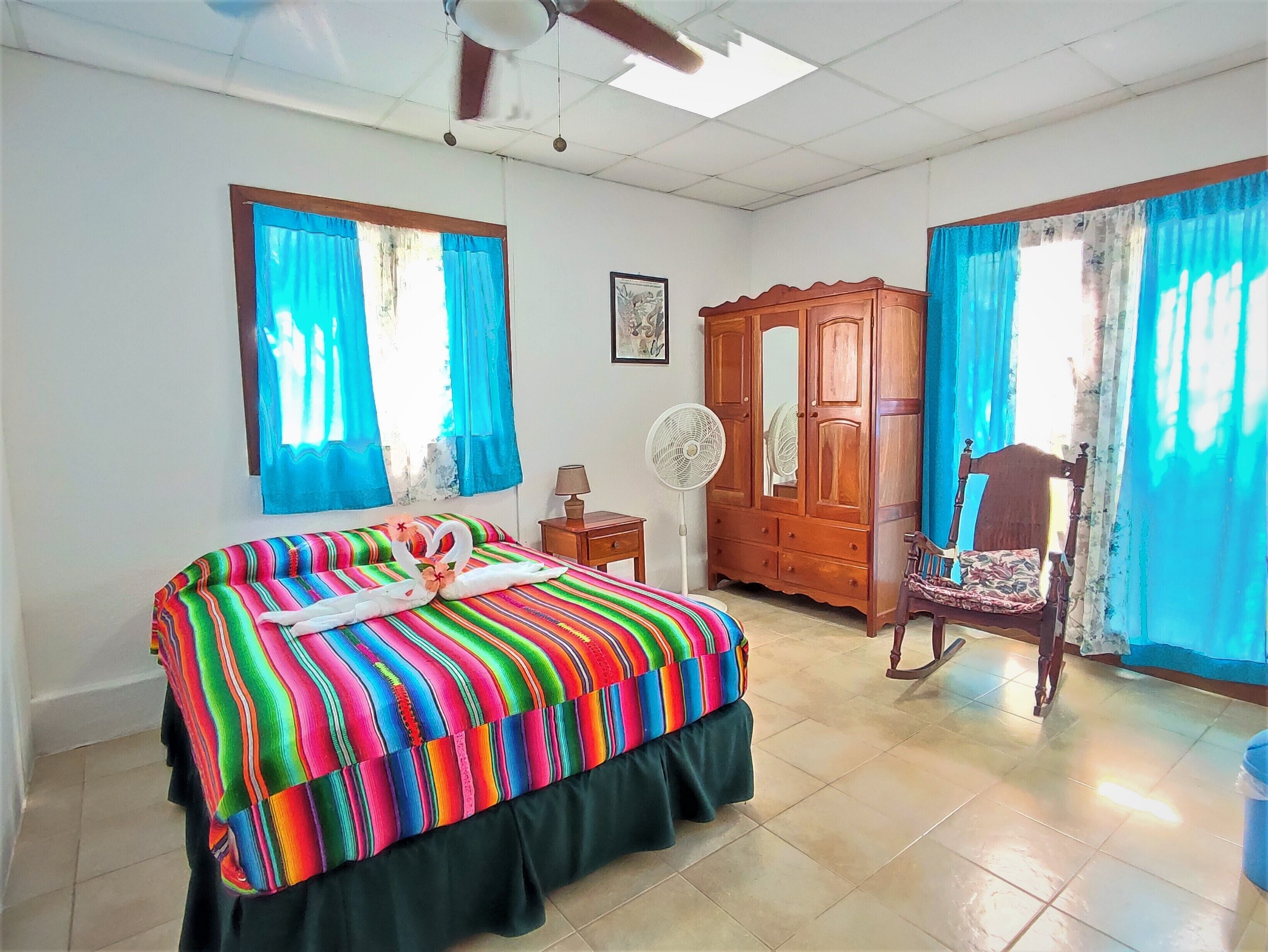Quarto com cama, guarda-roupa de madeira, cadeira, ventilador e ventoinha no teto.