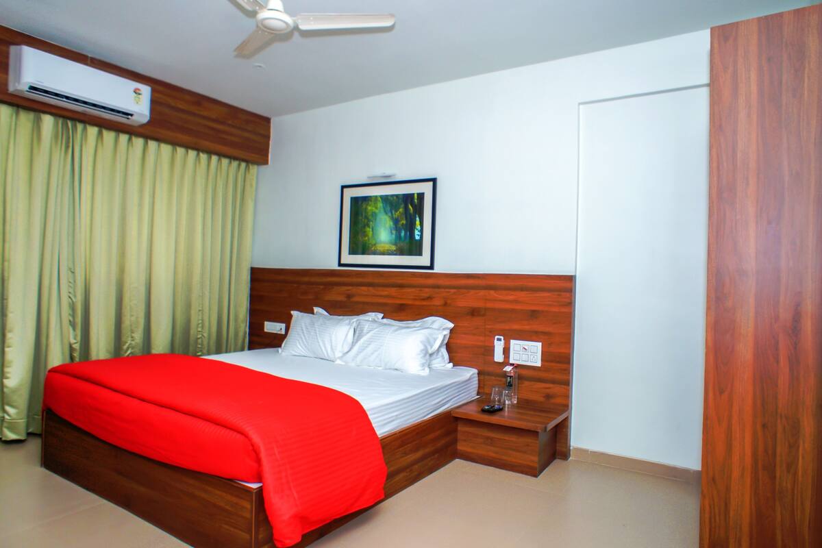 Quarto de hotel com cabeceira de madeira, cama com roupa de cama branca e manta vermelha, mesa de cabeceira com luminária, unidade de ar condicionado, quadro na parede e porta com painel de madeira.