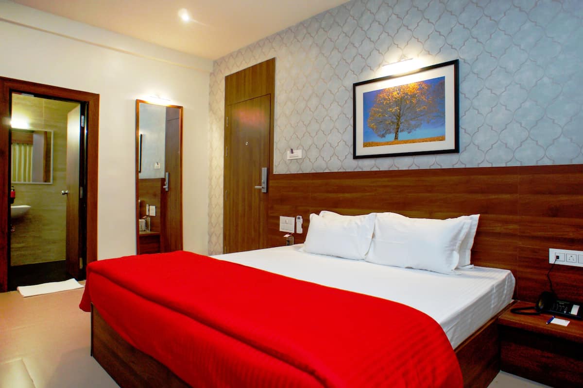 Quarto de hotel com uma cama grande, cabeceira de madeira, pintura de uma árvore, mesa de cabeceira com telefone e colcha vermelha.