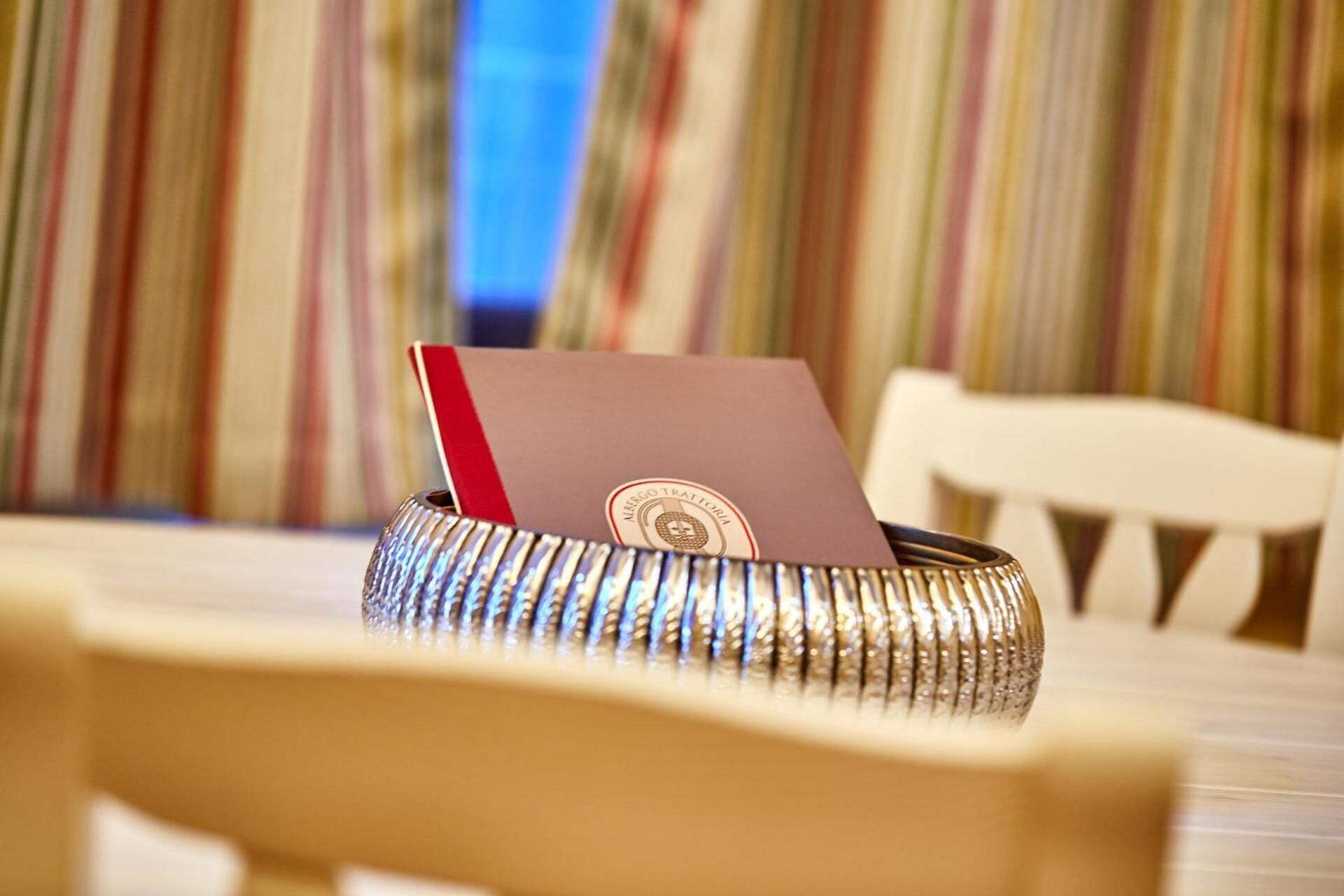 Um livro com capa vermelha colocado sobre um objeto metálico espiralado, possivelmente um item decorativo, em um quarto com cortinas listradas e uma cadeira branca.