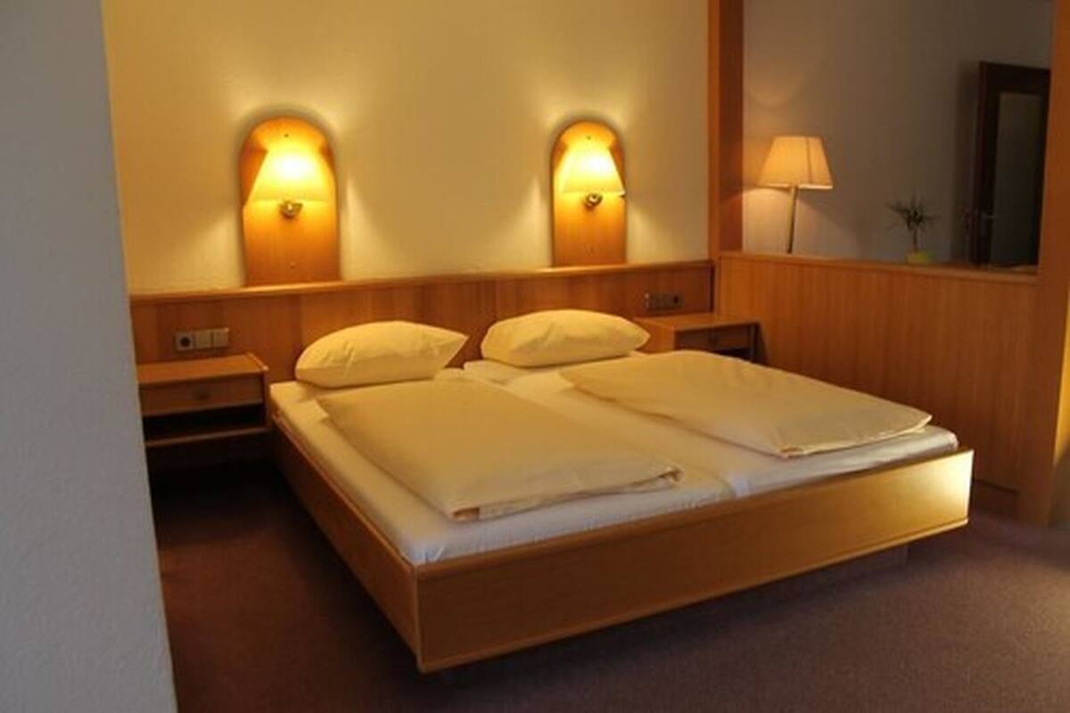 Uma cama de casal bem arrumada, com cabeceira de madeira, duas mesas de cabeceira e luminárias fixadas na parede.