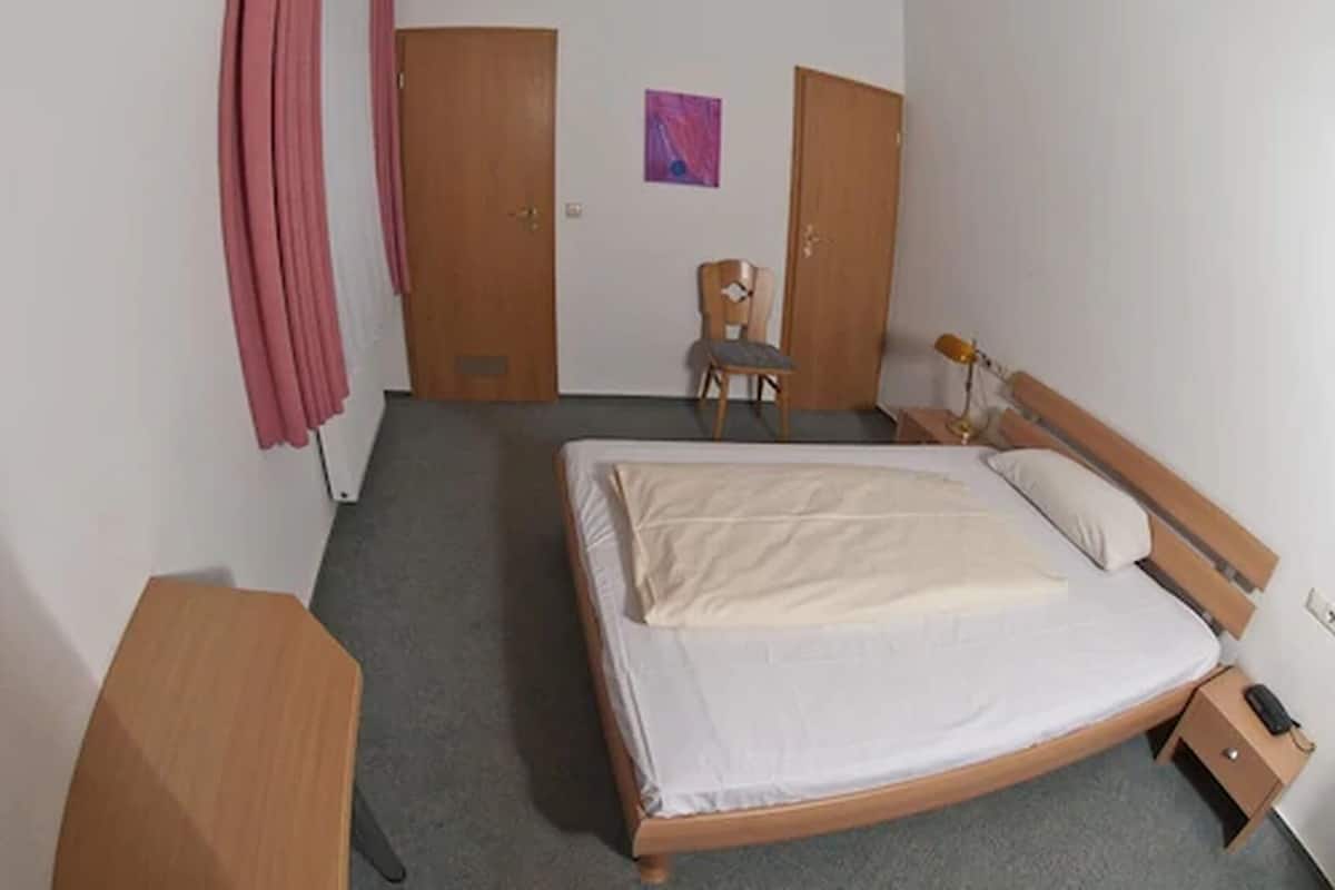 Quarto com uma cama de solteiro, uma mesa de cabeceira de madeira, uma cadeira e uma escrivaninha.