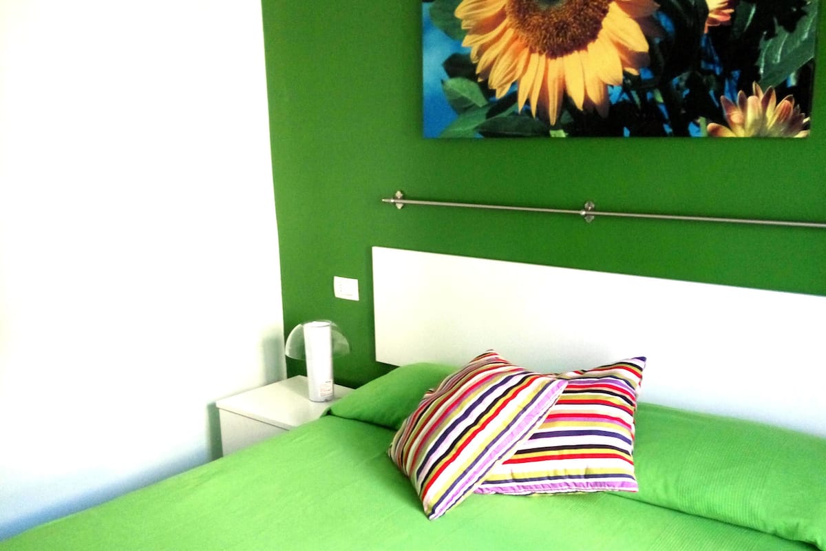 Uma cama com roupa de cama verde, um travesseiro listrado e colorido e um quadro de girassóis na parede.