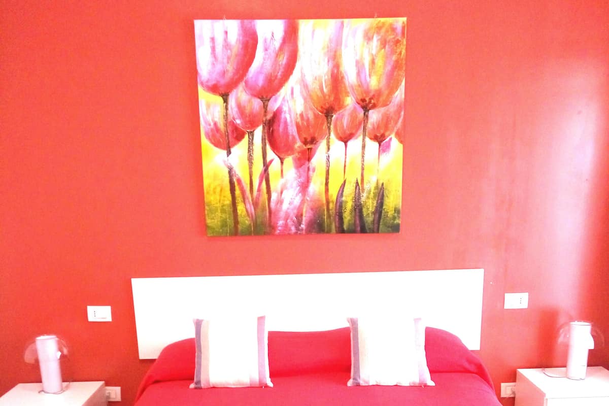 Quarto com colcha vermelha, cabeceira branca e duas mesas de cabeceira com luminárias.