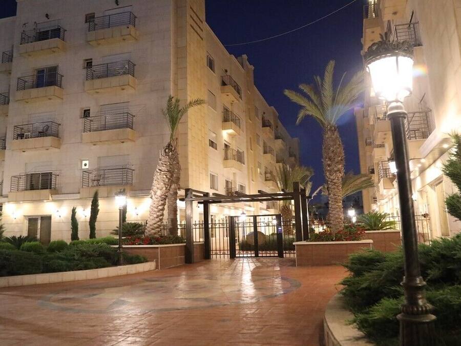 Um hotel com vários andares, entrada fechada por portão, palmeiras e um caminho bem iluminado.