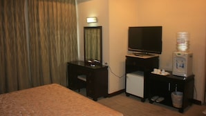 Quarto de hotel com cama, escrivaninha, televisão, pequeno frigorífico, dispensador de água e espelho.