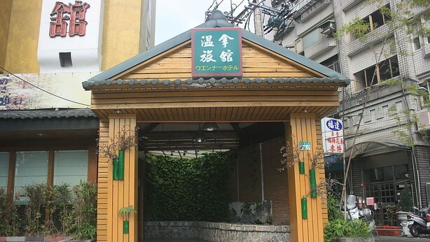 Uma entrada em estilo japonês tradicional, com estrutura de madeira, placa indicando recepção calorosa e uma pequena área de jardim com plantas.