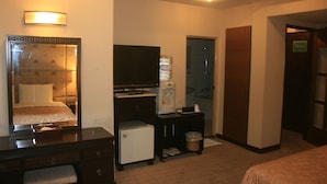 Quarto de hotel com cama, escrivaninha com espelho, televisão, mini-geladeira, dispensador de água e lixeira.