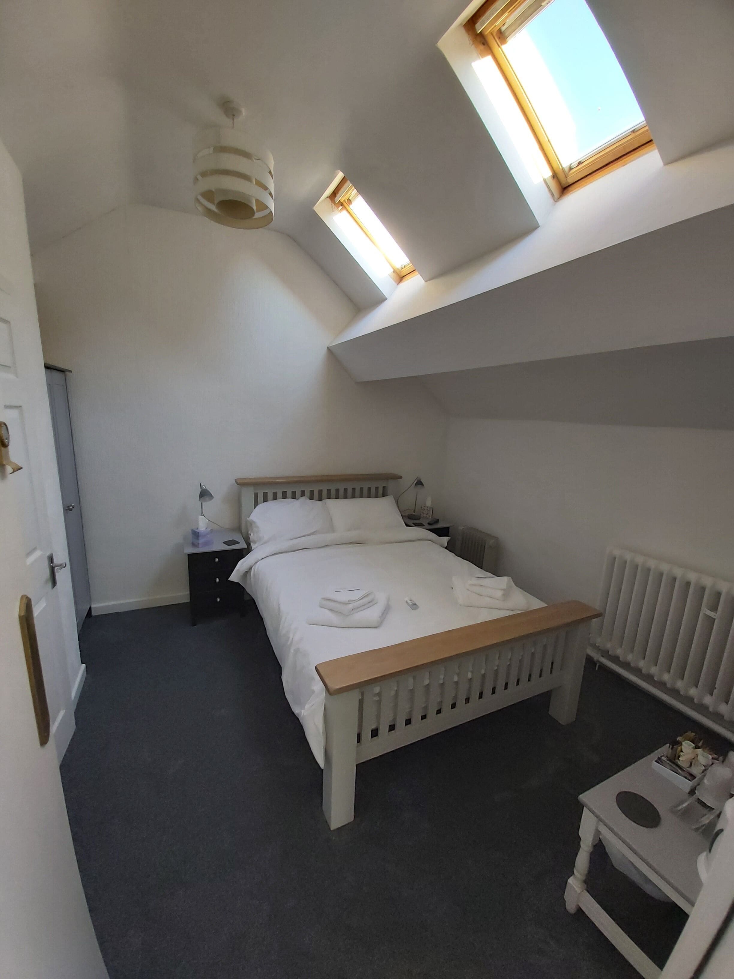 Quarto com claraboia, cama, mesa de cabeceira e radiador.