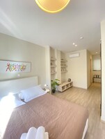 Quarto de hotel moderno com uma cama grande, uma mesinha de cabeceira com luminária, uma pequena escrivaninha com cadeira, uma planta em vaso e um ar-condicionado fixado na parede.