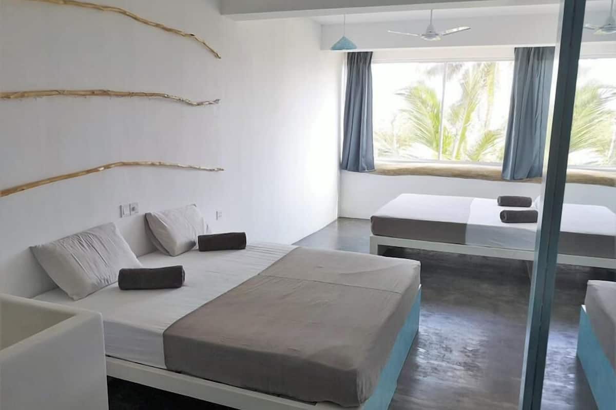 Quarto de hotel moderno com uma cama grande, uma cama menor e um sofá. Há duas janelas com cortinas e vista para palmeiras lá fora.