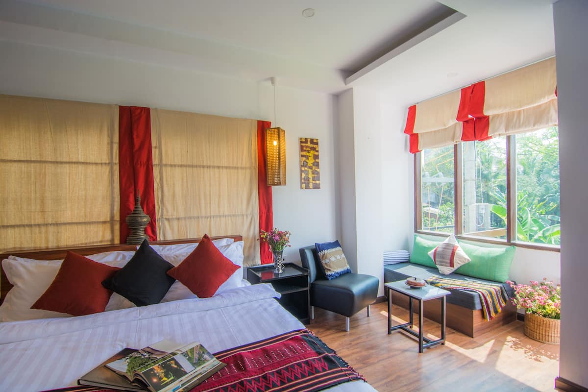 Quarto bem iluminado com uma cama grande, um espaço de estar com sofá e poltrona, e uma janela com vista para vegetação.