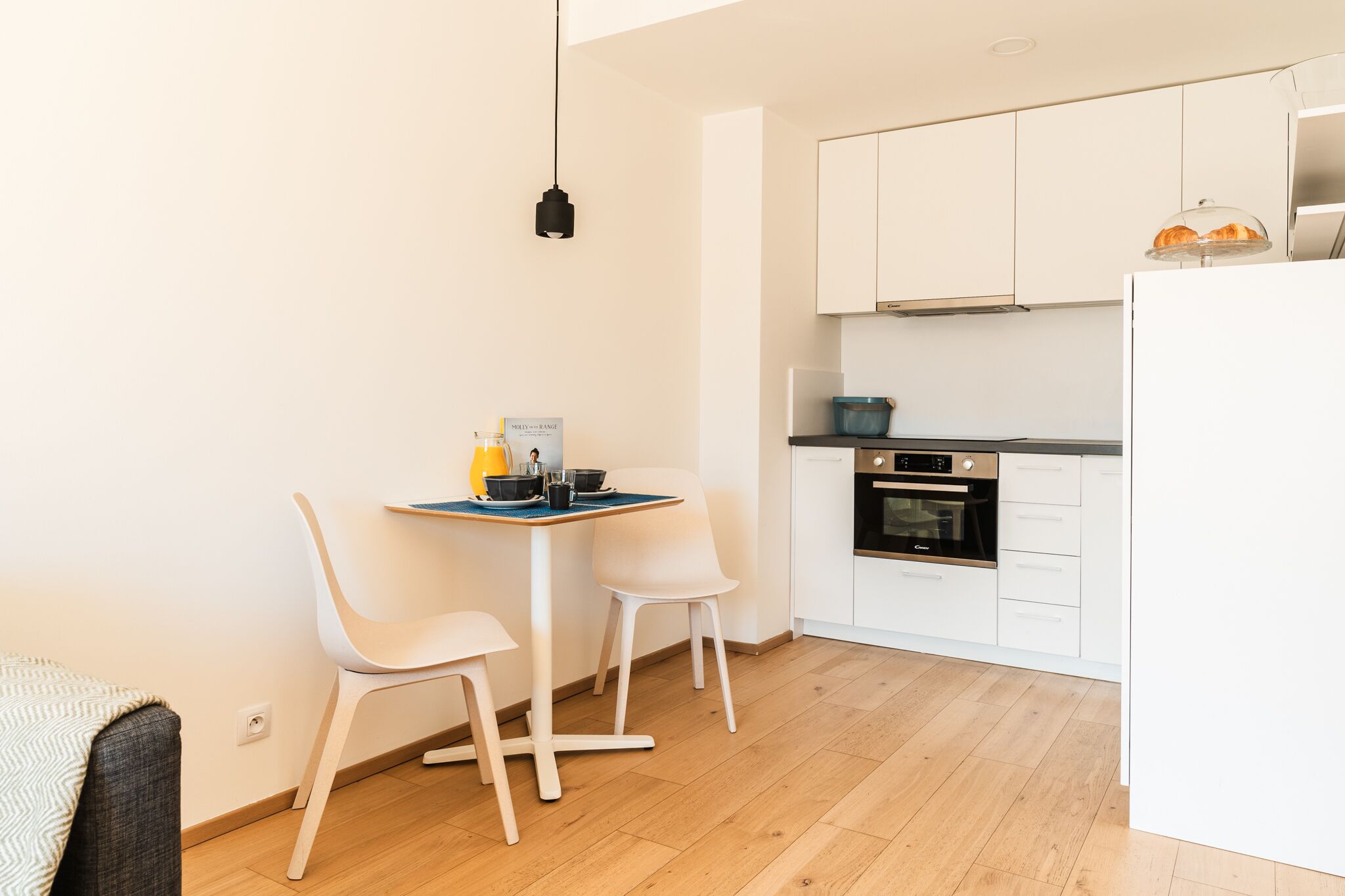 Uma cozinha compacta com armários brancos, forno preto e uma área de refeições com duas cadeiras brancas e uma mesa pequena.