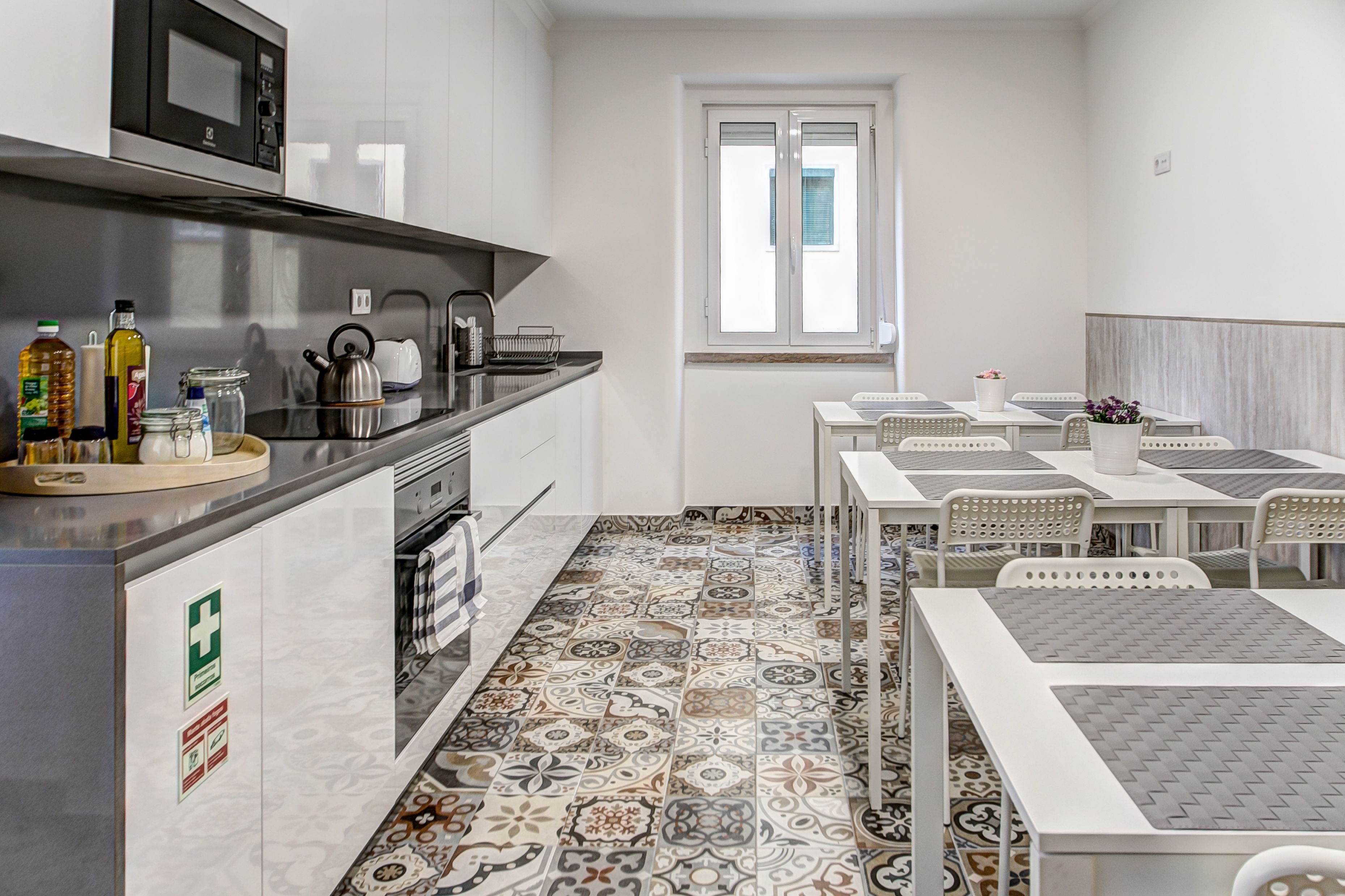 Cozinha compacta com micro-ondas, chaleira e diversos utensílios sobre o balcão. Possui um espaço de refeições com mesa e cadeiras, e uma janela com persianas.