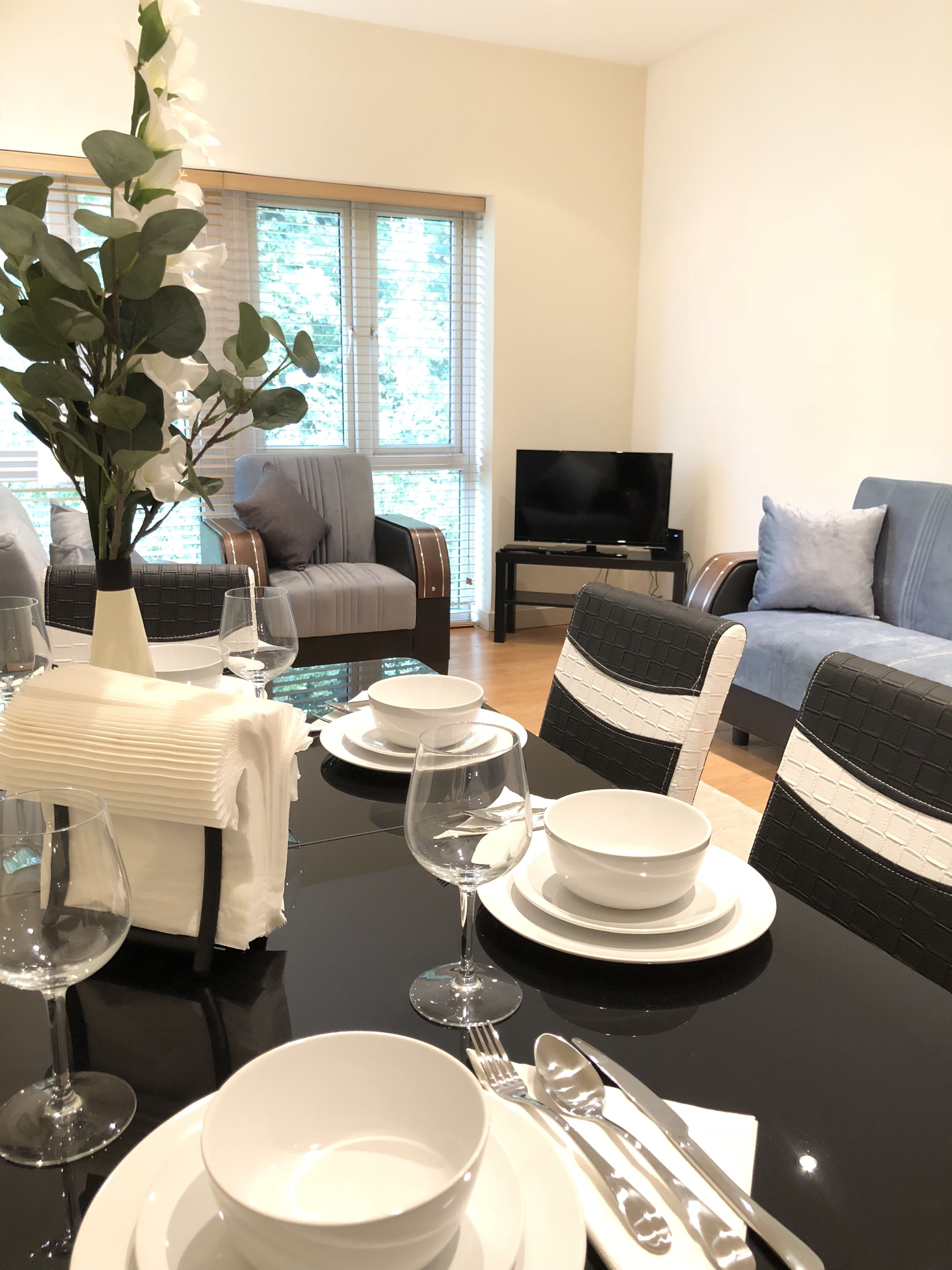 Uma sala de jantar com uma mesa preta posta para uma refeição, incluindo pratos, tigelas, copos de vinho e talheres. Há uma televisão ao fundo e um sofá com almofadas listradas.