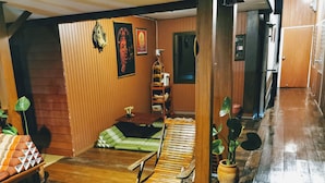 Quarto com piso de madeira, sofá com almofadas estampadas, um banco, prateleira com objetos decorativos e um vaso de planta.