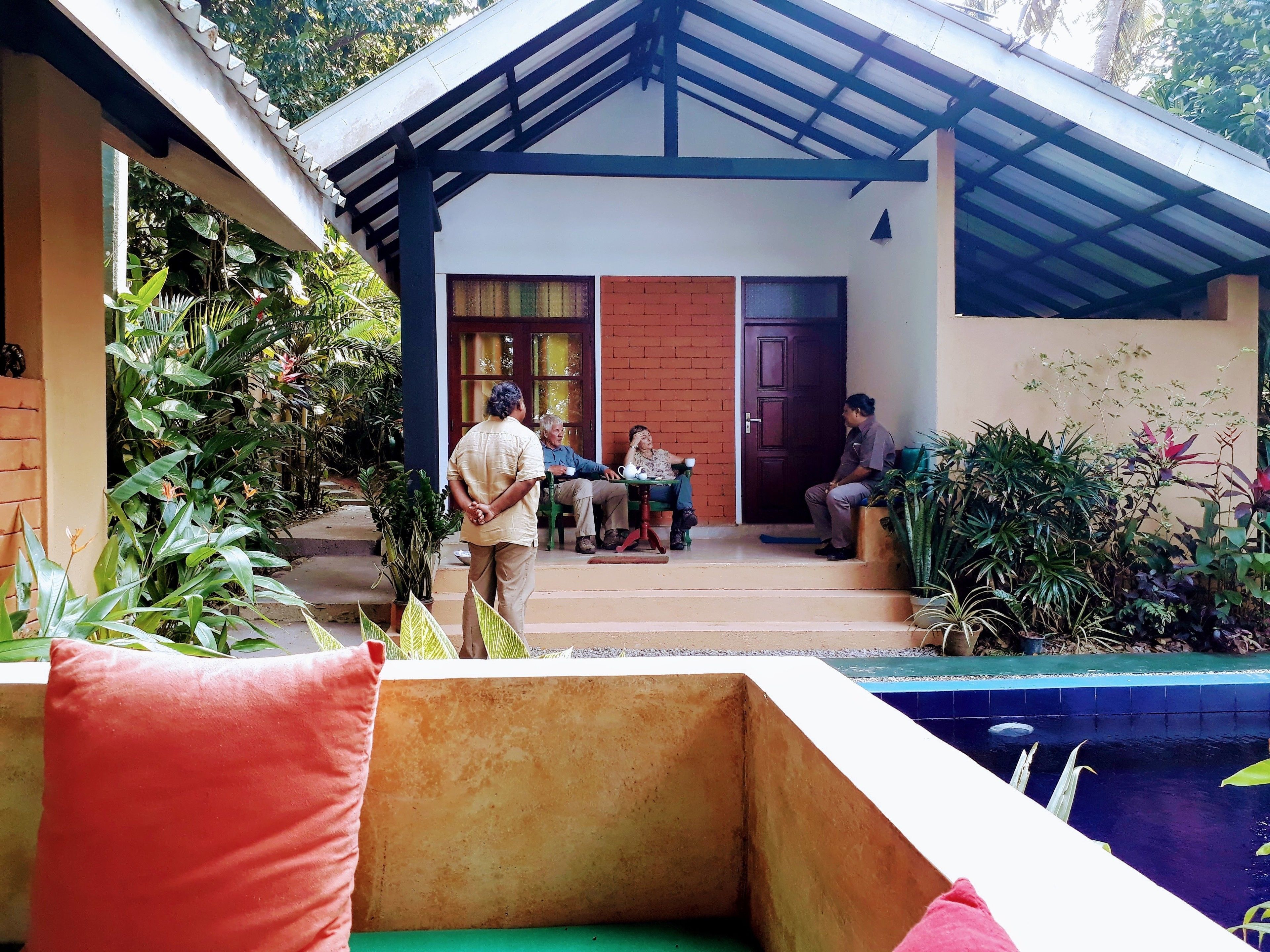 Uma área de estar coberta ao ar livre com piscina, rodeada por plantas e um muro de tijolos.