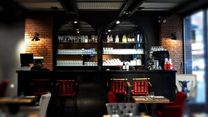 Uma área de bar com paredes de tijolo, prateleiras cheias de garrafas, copos e um balcão. Há cadeiras e mesas vermelhas, com alguns clientes sentados.