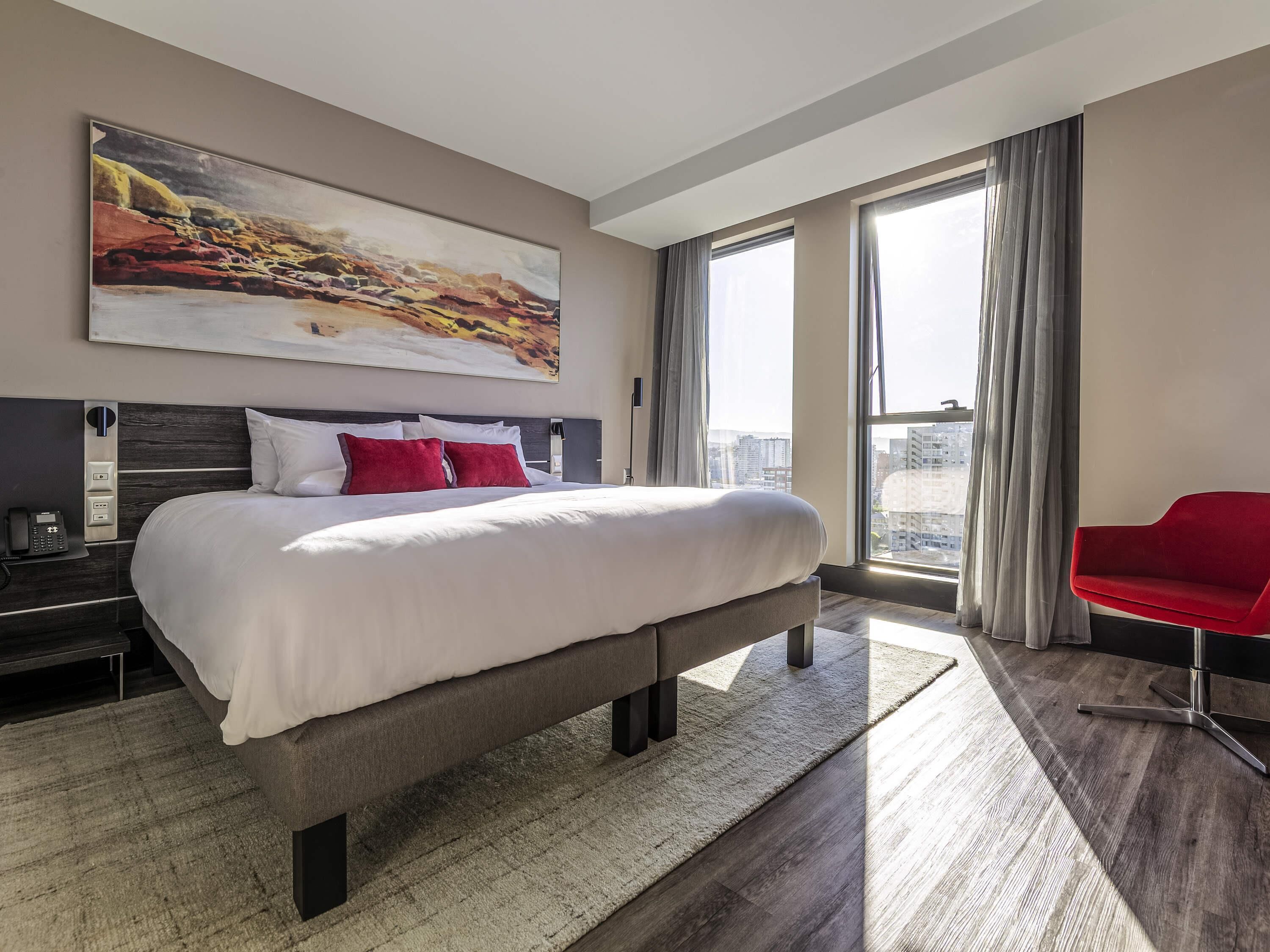 Quarto de hotel moderno com cama grande, uma cadeira vermelha, um quadro na parede e vista para a cidade.
