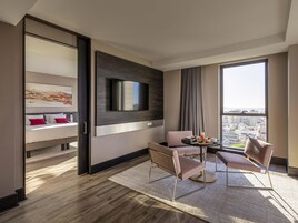 Quarto de hotel moderno com cama, TV fixada na parede, área de refeições com mesa e cadeiras, e uma grande janela com vista para a cidade.