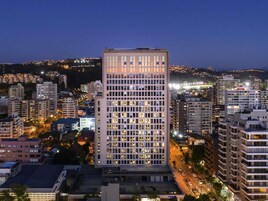 Um edifício alto com uma fachada em grade de janelas, cercado por outros prédios e uma paisagem urbana ao entardecer.