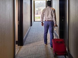 Um homem caminha por um corredor com uma mala vermelha, em direção a uma porta com vista para edifícios lá fora.