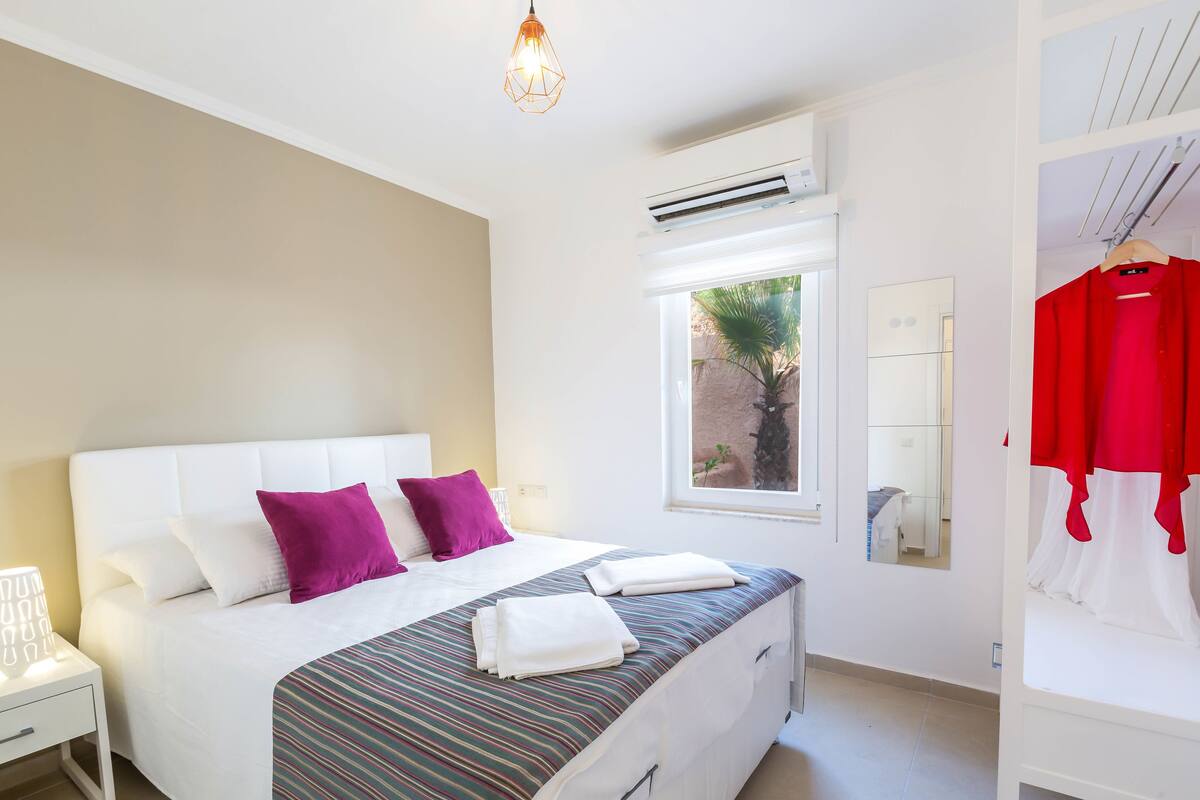 Quarto com cama, mesas de cabeceira brancas, janela com vista, aparelho de ar condicionado e uma peça de roupa vermelha em um cabide.
