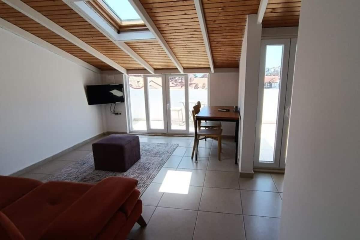 Quarto com teto de madeira, televisão na parede, mesa de jantar com cadeiras e uma janela grande.