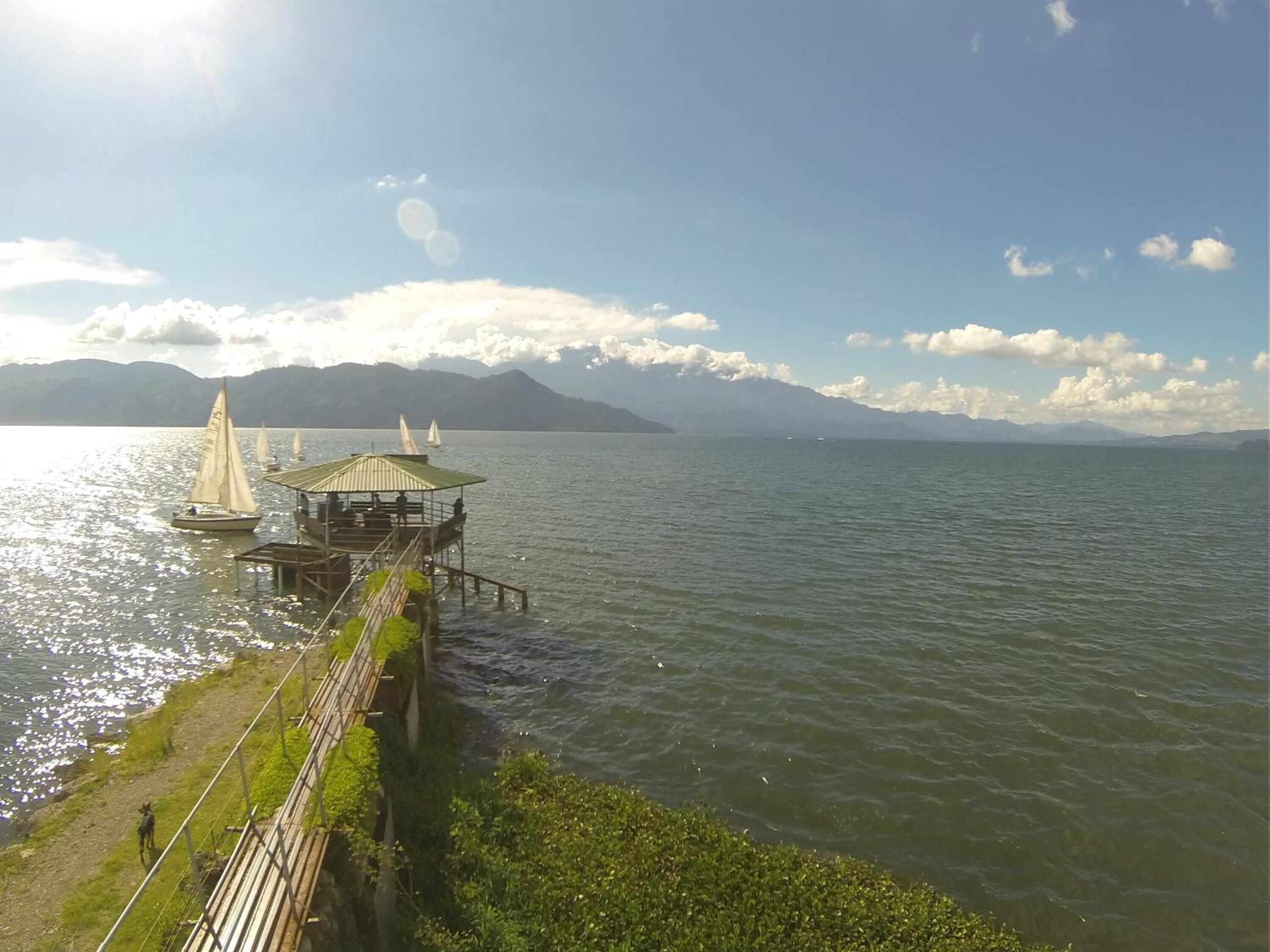 Um píer se estende sobre um lago, com um coreto e um veleiro.