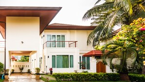 Uma casa de dois andares com varanda, entrada coberta e um jardim bem cuidado com palmeiras e plantas em vasos.