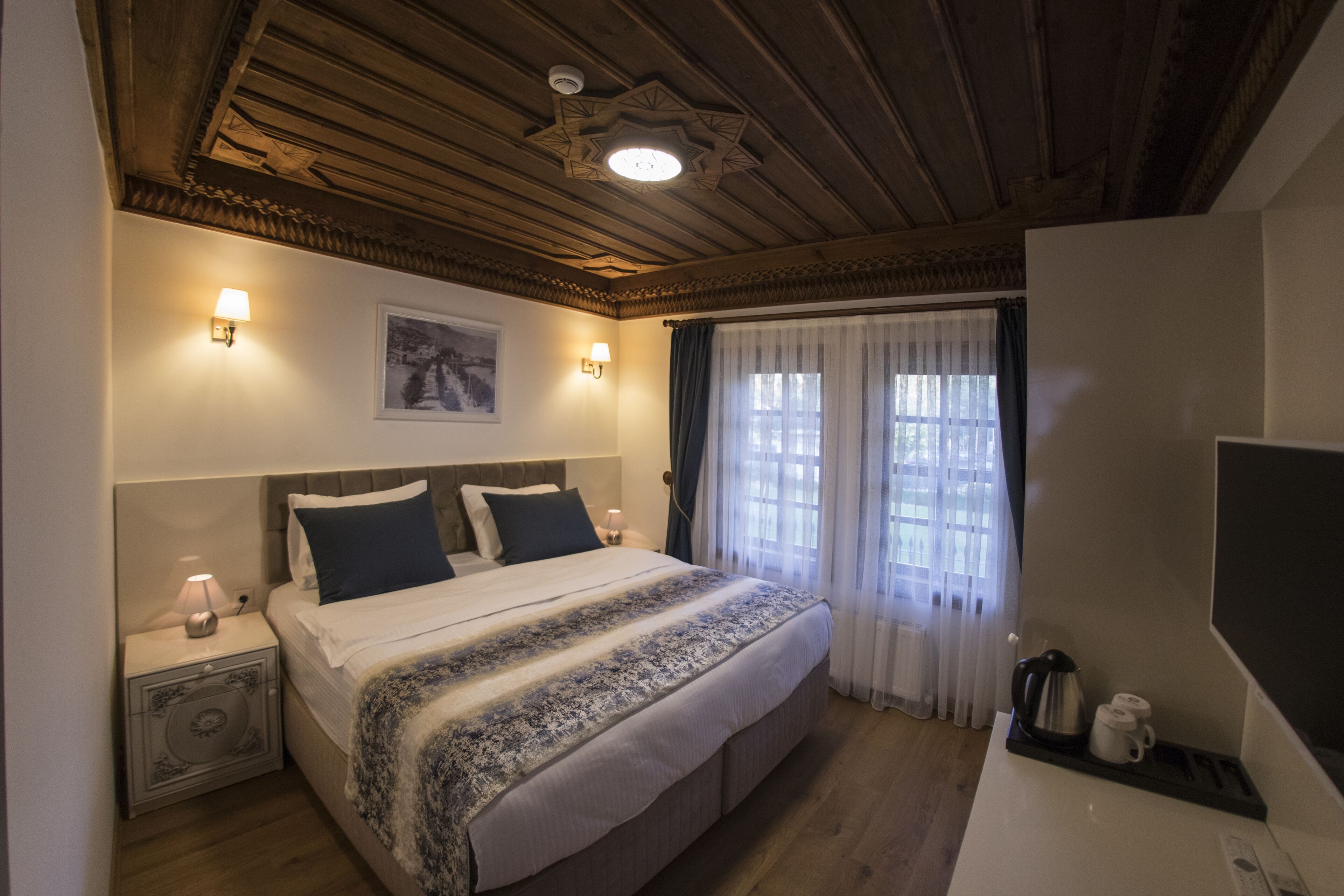 Quarto com cama grande, duas luminárias de cabeceira, luz de teto, janela com cortinas e uma máquina de café em uma mesinha.