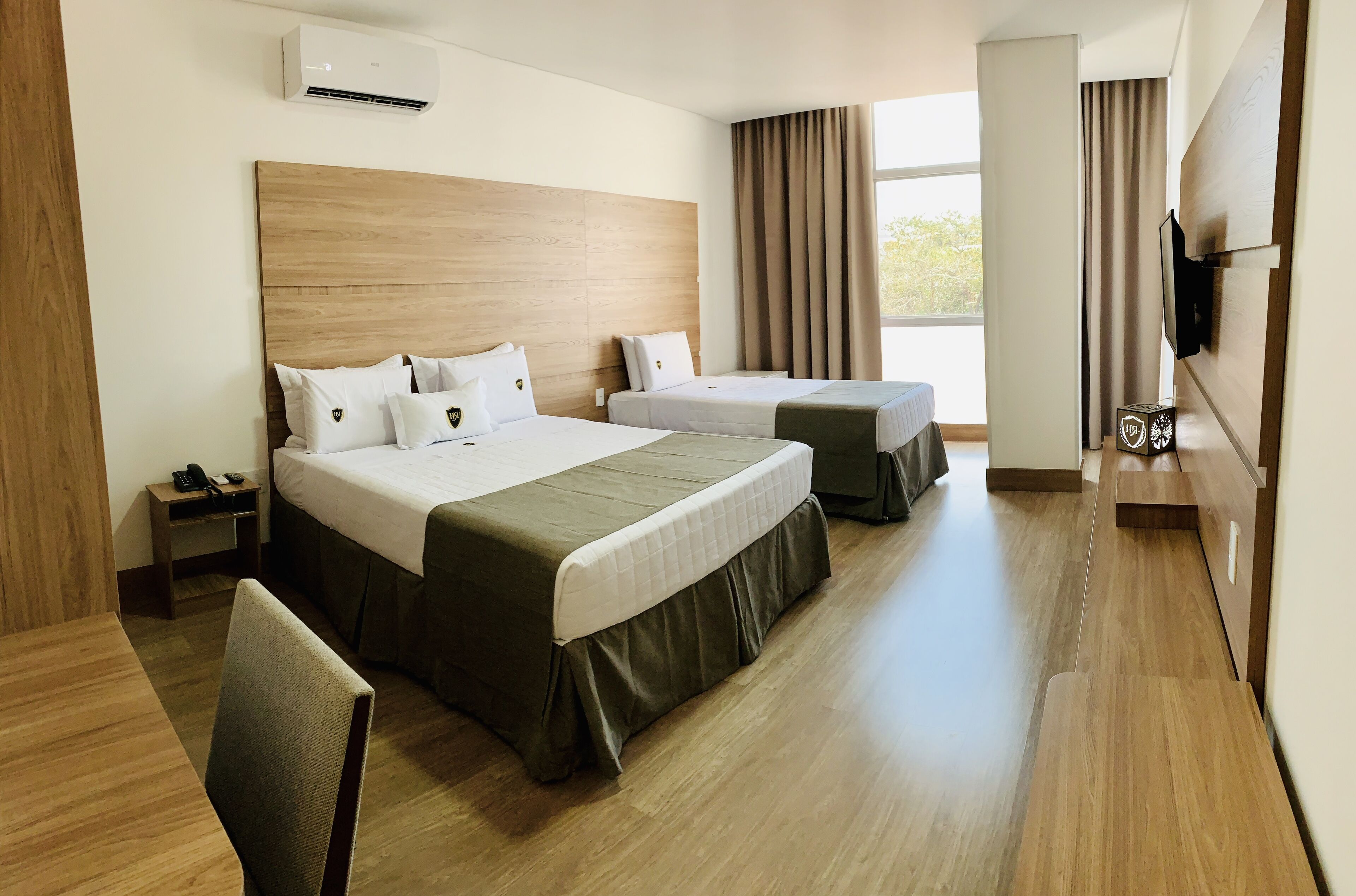 Quarto de hotel com duas camas, uma escrivaninha, uma cadeira, uma televisão e uma janela com cortinas.