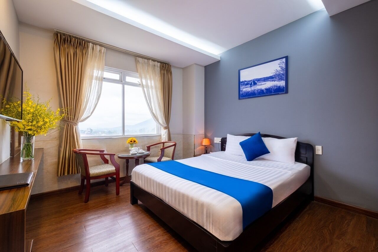 Quarto de hotel com cama, escrivaninha, cadeira, pequena mesa com um vaso de flores e janela com cortinas.