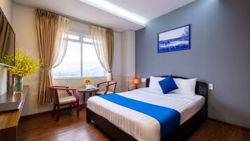 Quarto de hotel com cama, escrivaninha, cadeira, pequena mesa com um vaso de flores e janela com cortinas.