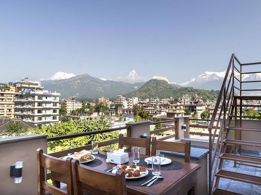 Uma área de refeições com uma mesa posta para dois, com vista para uma paisagem montanhosa e edifícios ao fundo.