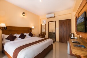 Quarto de hotel com uma cama grande, cabeceira de madeira, televisão fixada na parede, escrivaninha com chaleira e porta para o banheiro.