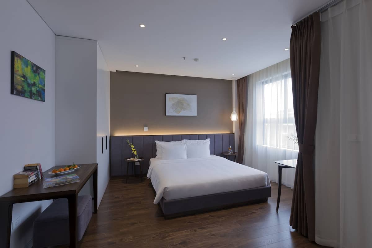 Quarto de hotel moderno com uma cama grande, uma mesinha de cabeceira, uma escrivaninha e uma janela com cortinas.