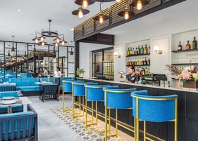 Uma área de bar moderna com banquetas azuis, um bartender preparando bebidas e diversas garrafas em prateleiras.
