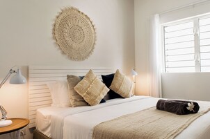 Uma cama bem arrumada com cabeceira branca, um painel de parede tecido e uma janela com persianas.