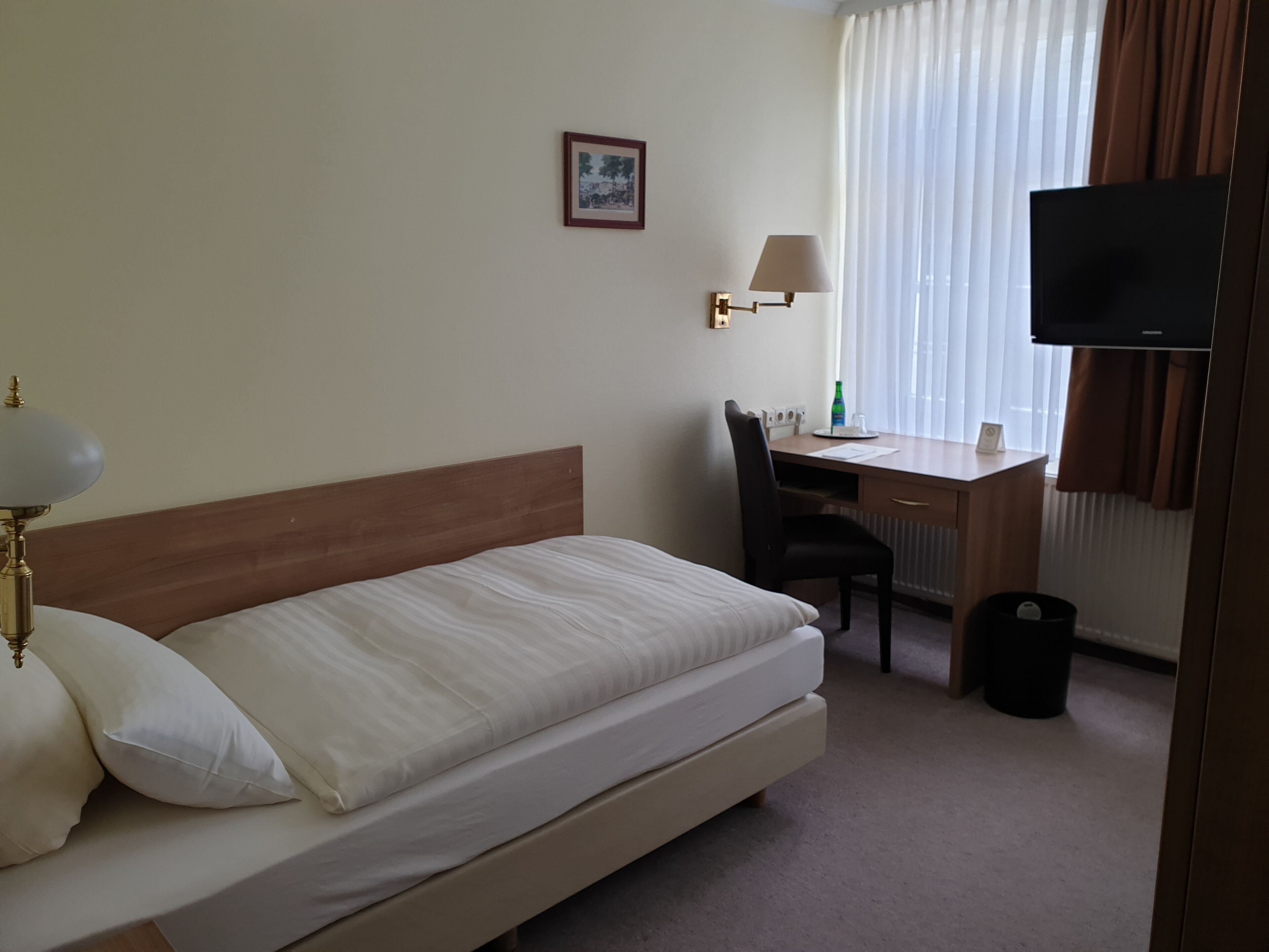 Quarto de hotel com cama, escrivaninha, cadeira, luminária, televisão e um quadro na parede.
