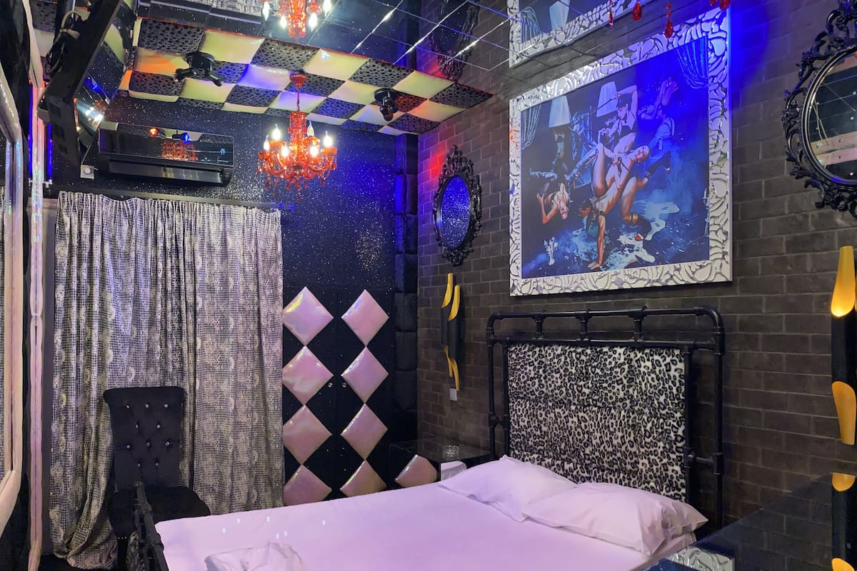 Quarto com cama, um lustre, um espelho grande e televisão na parede.