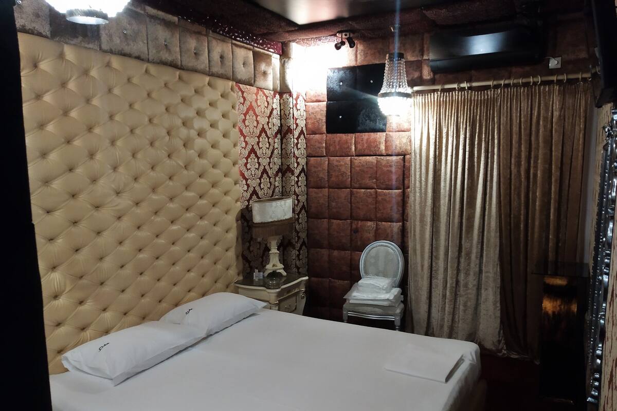 Quarto com cabeceira estofada, mesa de cabeceira com abajur e janela com cortinas.