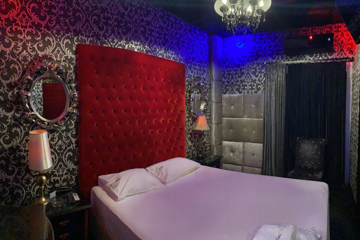 Quarto de hotel com cabeceira estofada vermelha, cama branca, espelho e lustre.