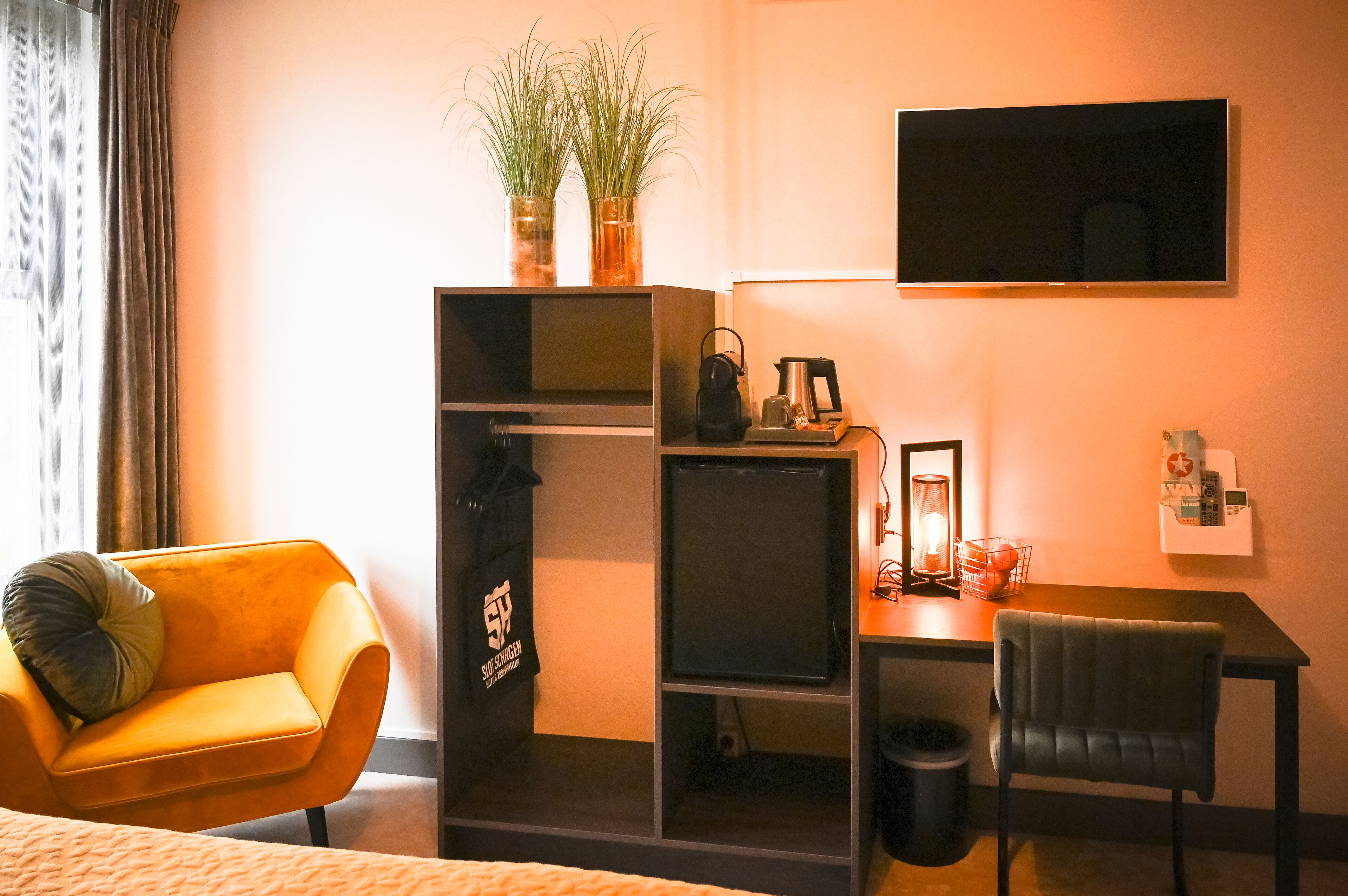 Quarto compacto com sofá, escrivaninha, cadeira, televisão, prateleira com plantas e lixeira.