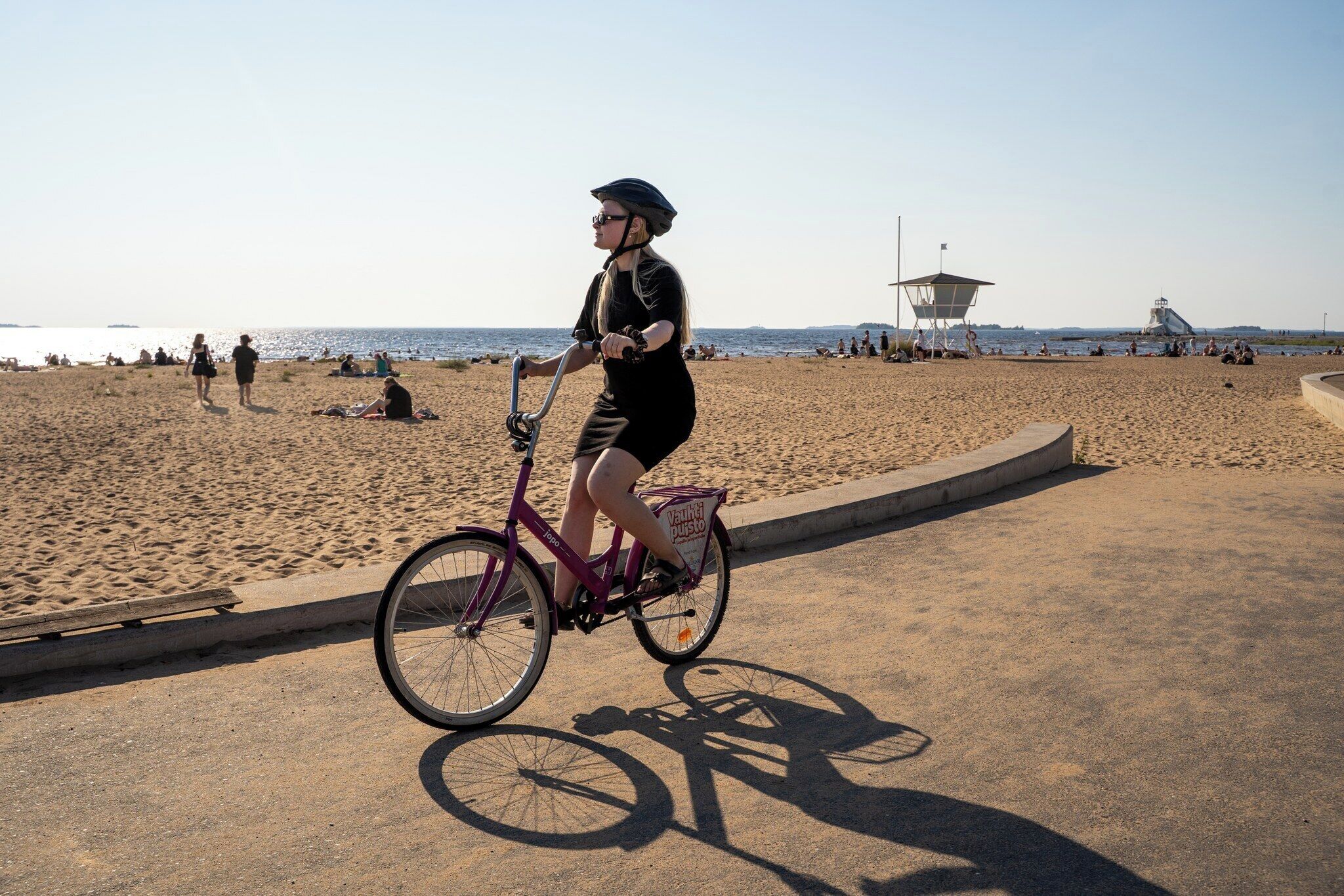 Uma pessoa andando de bicicleta em um caminho pavimentado perto de uma praia de areia, com uma torre salva-vidas ao fundo.