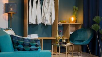 Quarto de hotel moderno com sofá verde-turquesa, mesa de vidro com garrafa e copos, cadeira verde-turquesa, cabideiro com roupões brancos e planta em vaso.
