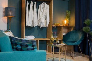 Quarto de hotel moderno com sofá verde-turquesa, mesa de vidro com garrafa e copos, cadeira verde-turquesa, cabideiro com roupões brancos e planta em vaso.